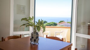 Uma mesa de madeira com uma planta em vaso, cadeiras brancas e vista para o mar através de uma grande janela.