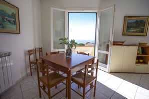 Uma sala de jantar com mesa e cadeiras de madeira, uma planta em vaso e vista para o mar por uma porta aberta.