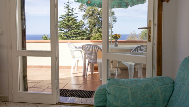 Quarto com porta de vidro deslizante que dá para um pátio externo com mesa e cadeiras, sofá verde com padrão e vista para o oceano.
