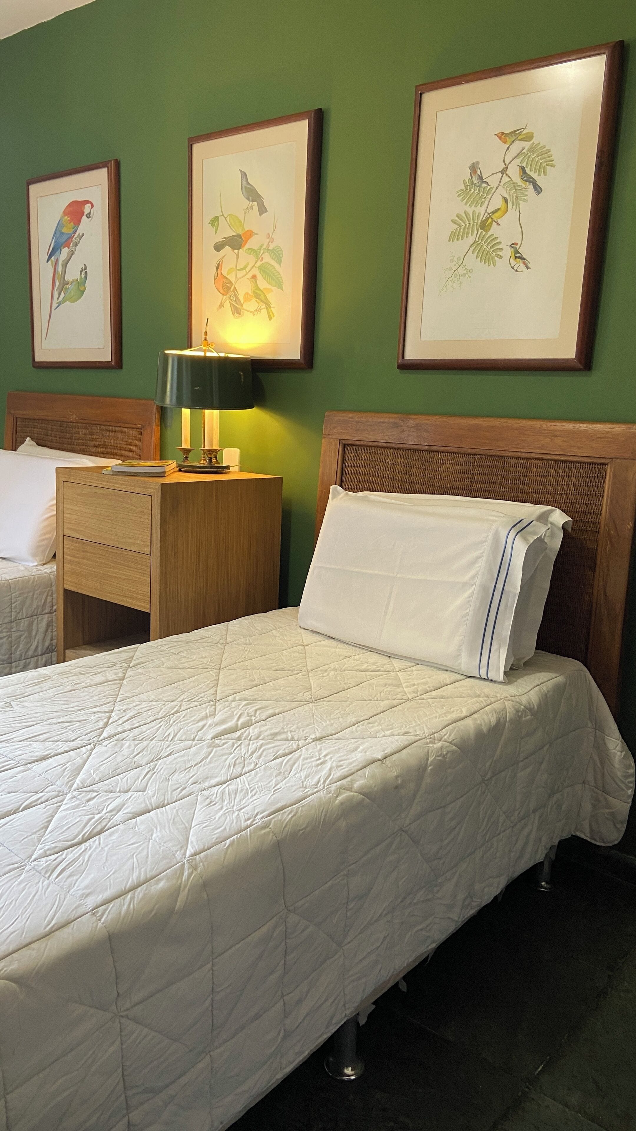 Quarto de hotel com uma cama grande, uma mesa de cabeceira com abajur, e três quadros de pássaros na parede.