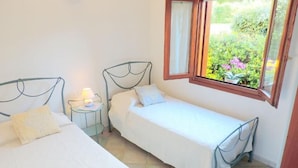 Quarto pequeno e limpo com duas camas de solteiro, uma mesa de cabeceira com abajur e uma janela com vista para o verde.