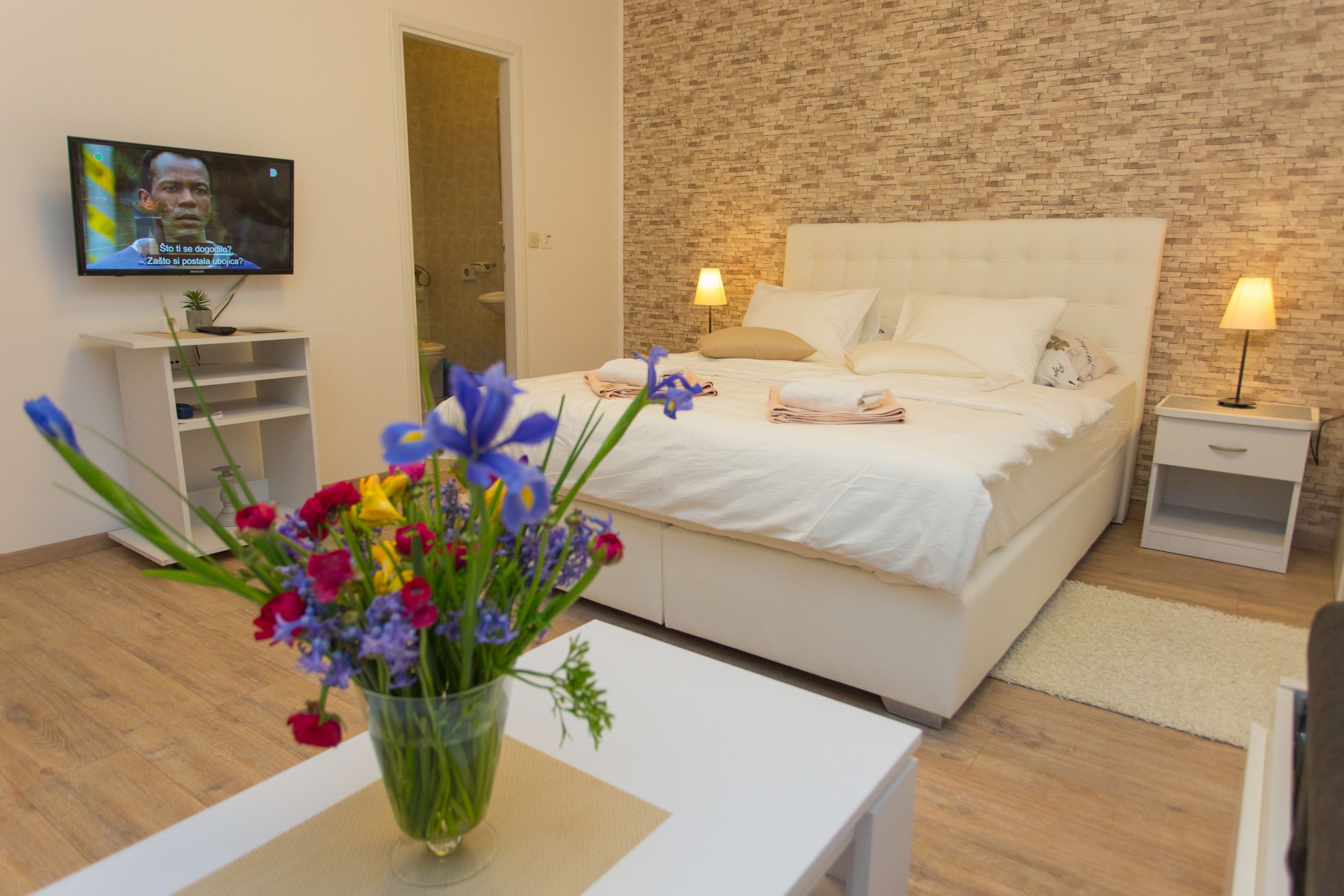 Quarto de hotel com uma cama grande, televisão fixada na parede, mesa de cabeceira com abajur, uma pequena planta e um vaso com flores.