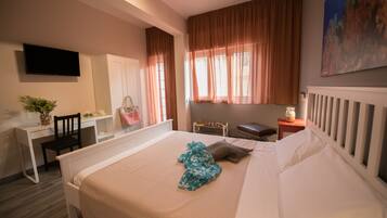 Quarto de hotel com cama, escrivaninha, cadeira, televisão e janela com cortinas.