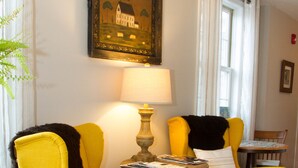 Quarto com duas poltronas amarelas, uma mesa pequena com abajur, um quadro emoldurado na parede e uma janela com cortinas.