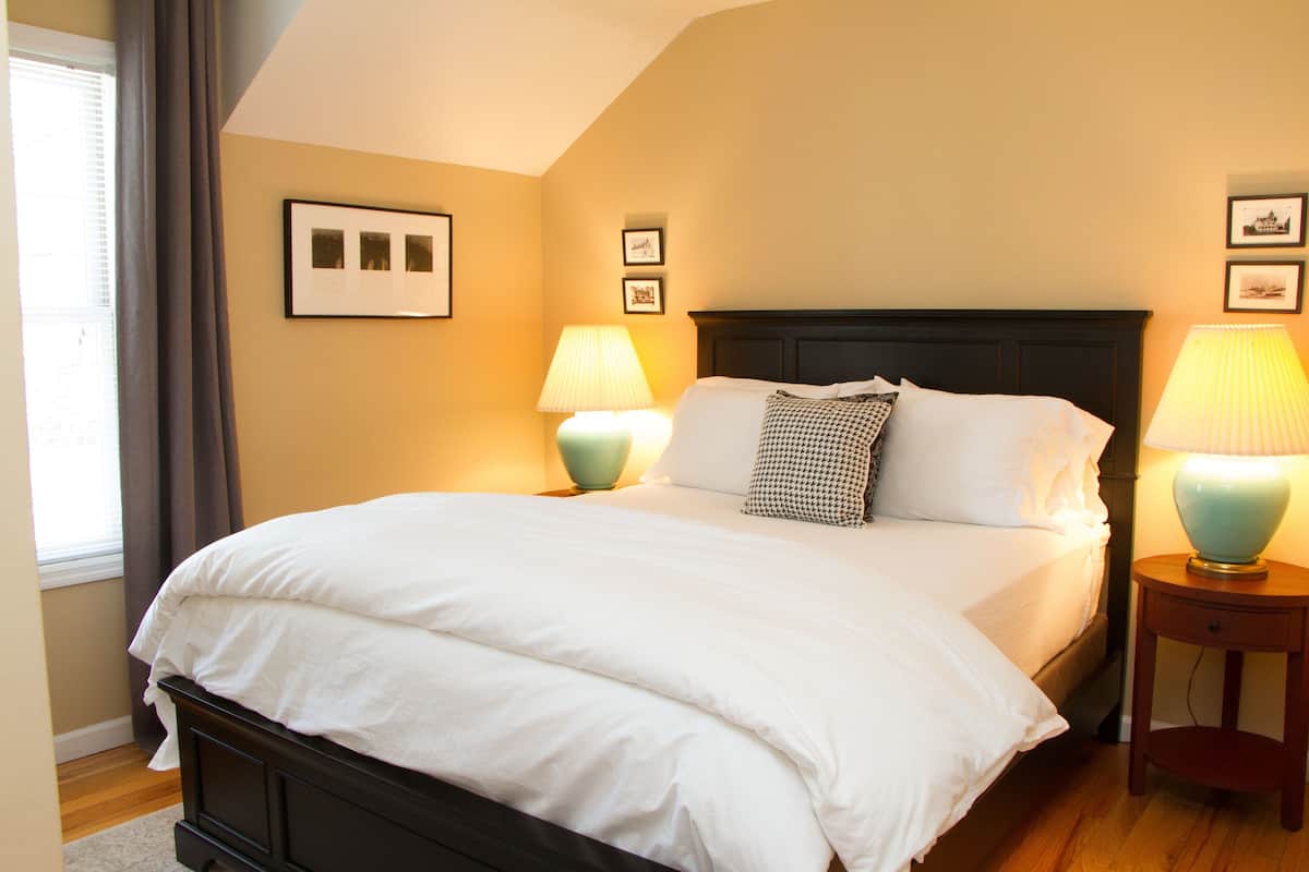 Uma cama bem arrumada com lençóis brancos, duas luminárias de cabeceira e um quadro na parede.