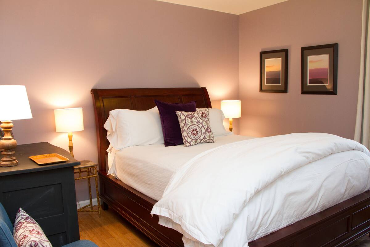 Uma cama bem arrumada com roupa de cama branca e roxa, cabeceira de madeira e duas luminárias fixadas na parede.