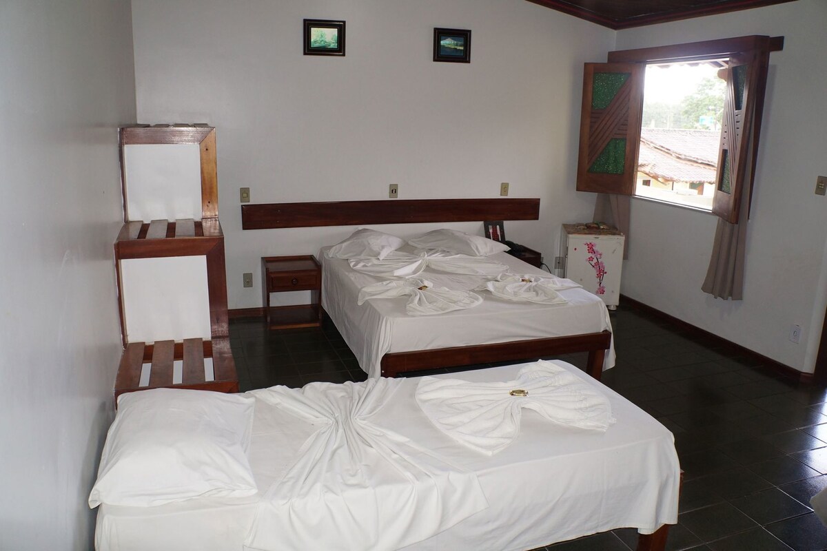 Quarto com cama, mesa de cabeceira, um banco de madeira, cômoda e janela com cortinas.