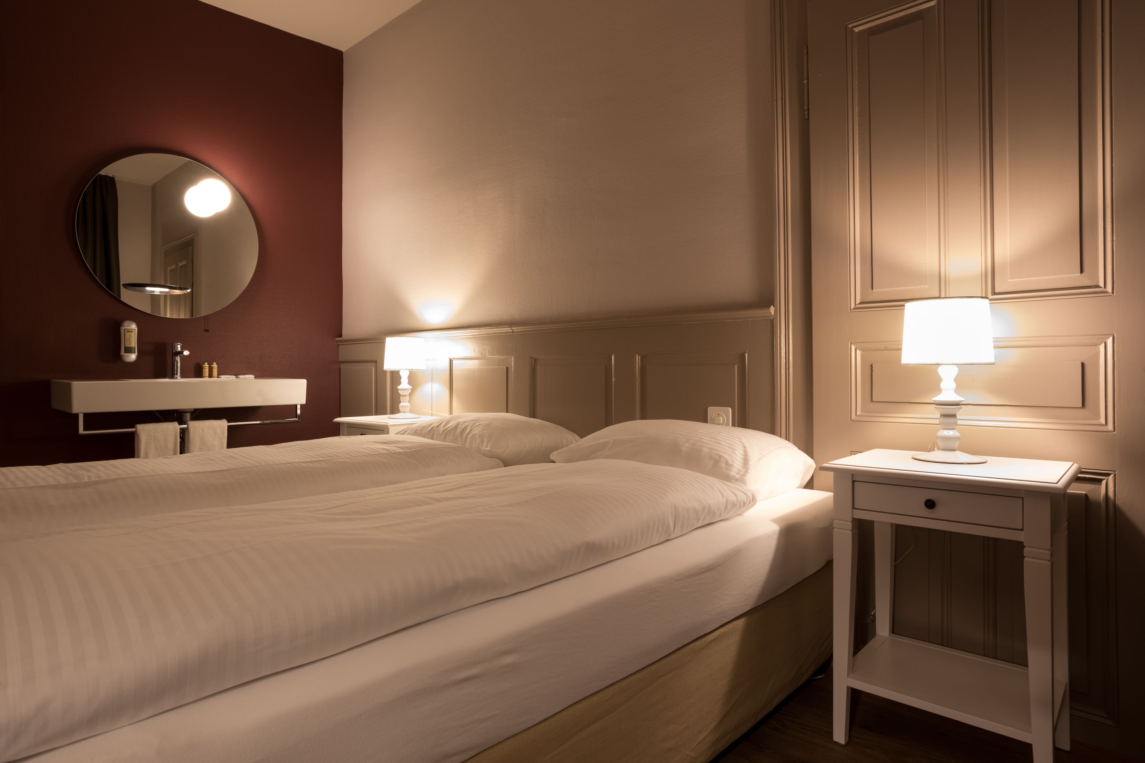 Quarto de hotel com cama grande, mesa de cabeceira com abajur, espelho redondo e pequeno armário.