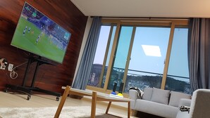 Sala de estar com televisão de tela plana, mesa de centro de madeira, sofá cinza e grandes janelas com vista para uma ponte.