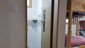 Corredor estreito que dá para um banheiro com chuveiro e vaso sanitário. No quarto adjacente, há uma cama com colchão rosa e cabeceira de madeira.