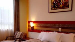 Quarto de hotel com uma cama grande, cabeceira com padrão decorativo, um quadro na parede e uma janela com cortinas.