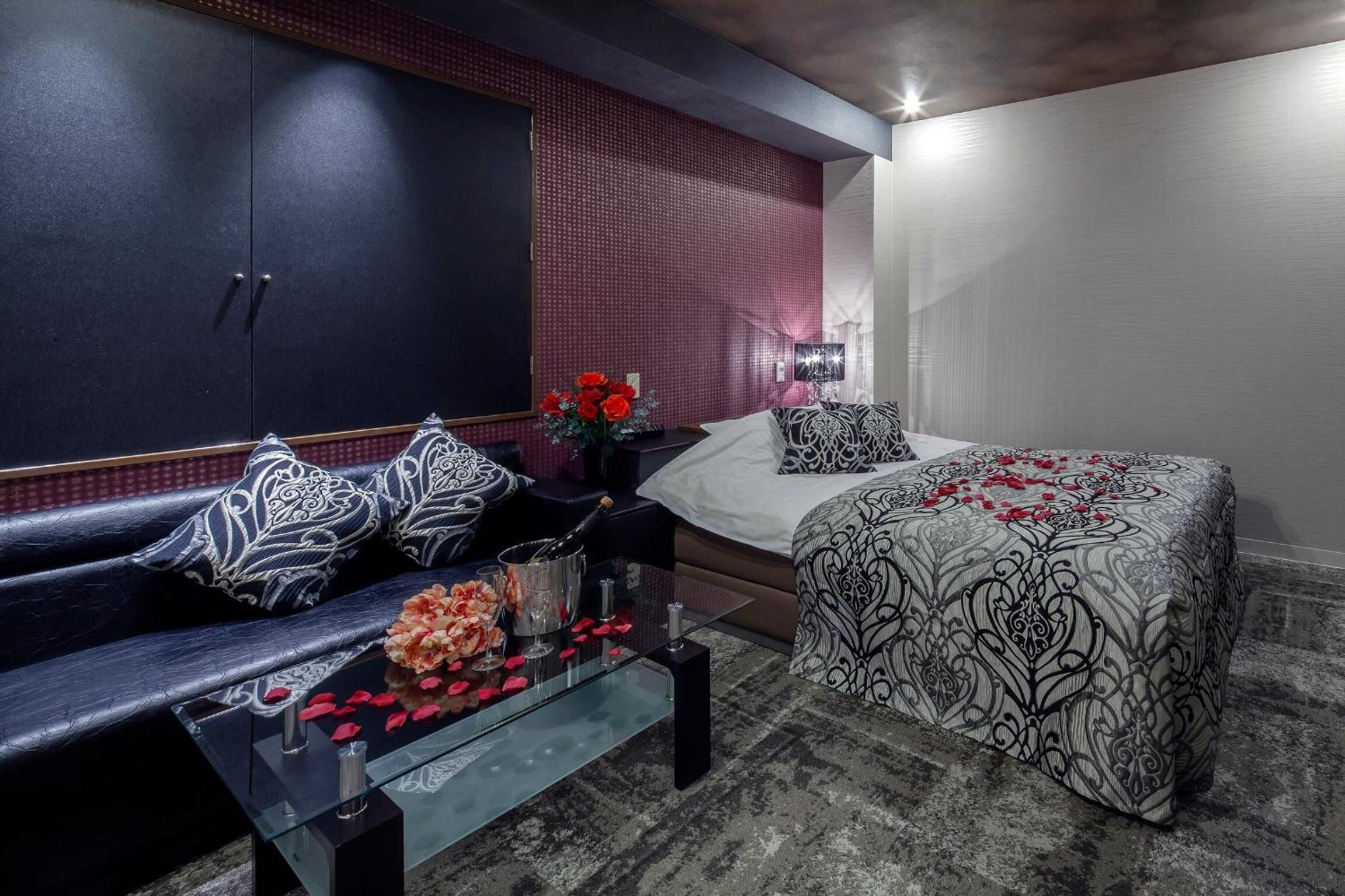 Quarto moderno com um sofá azul escuro, uma mesa de centro de vidro com um vaso de flores, uma cama com colcha estampada e uma parede vermelha com textura.