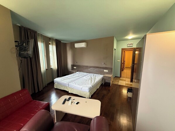 Quarto de hotel com cama, uma mesinha, uma cadeira vermelha, televisão e janela com cortinas.