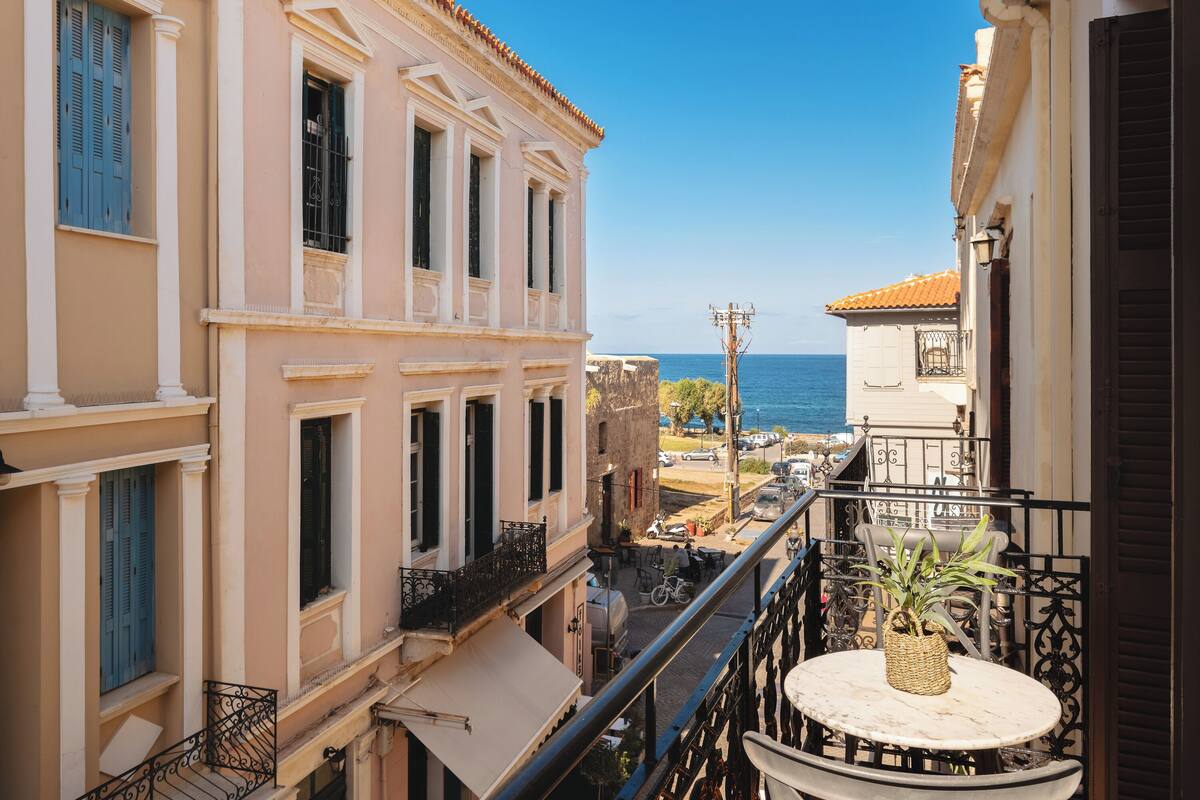 Uma varanda com vista para o mar, uma mesa redonda com uma planta e um edifício com persianas azuis.