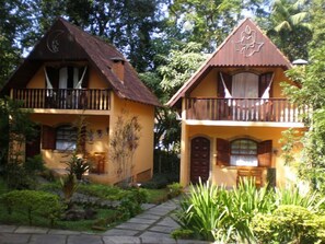 Duas pequenas casas de dois andares com varandas, cercadas por vegetação exuberante e um caminho de pedra.