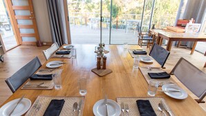 Uma sala de jantar com mesa de madeira posta para refeição, rodeada por cadeiras, e vista para o exterior.