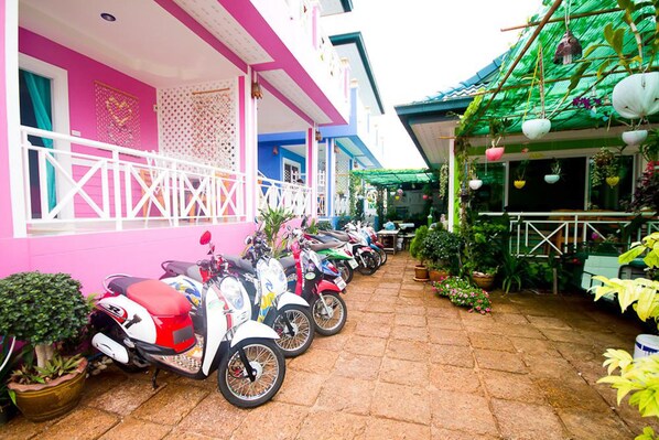 Fileira de scooters estacionadas em frente a um edifício com fachada rosa e azul, plantas em vasos e área de pátio com azulejos.