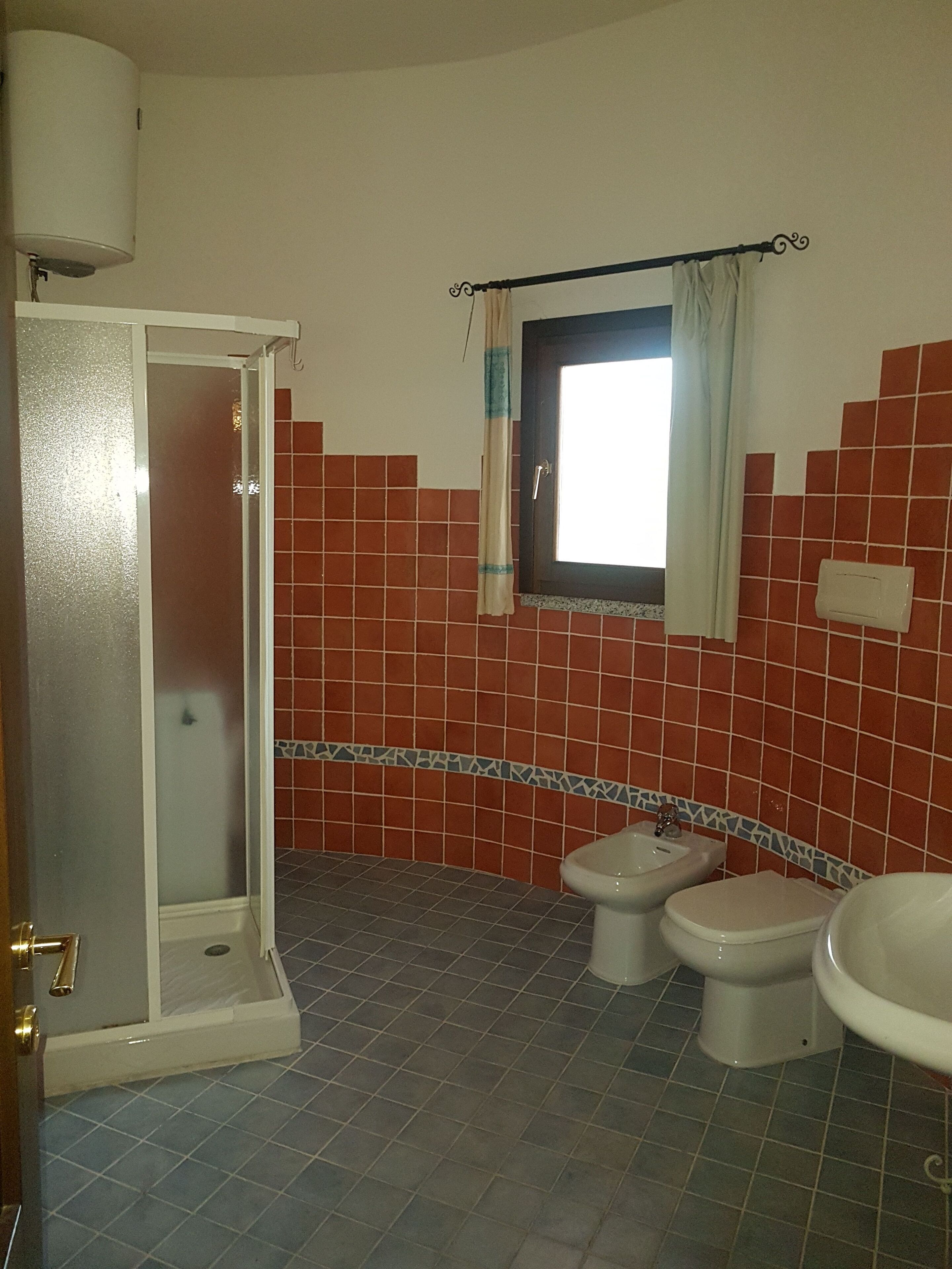 Um banheiro com chuveiro, dois vasos sanitários, pia e janela com cortinas.