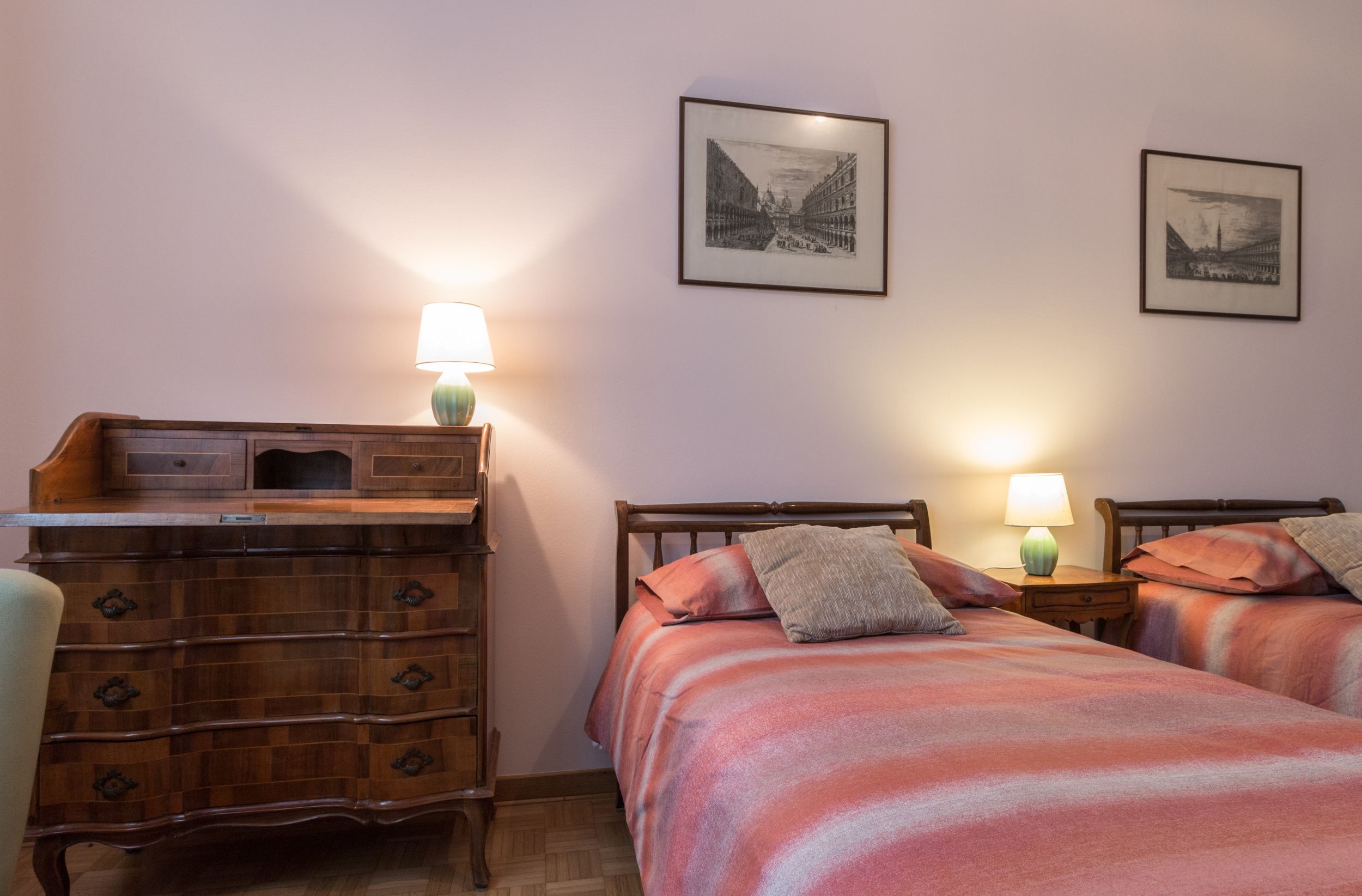 Quarto com duas camas, cômoda de madeira e duas luminárias fixadas na parede.