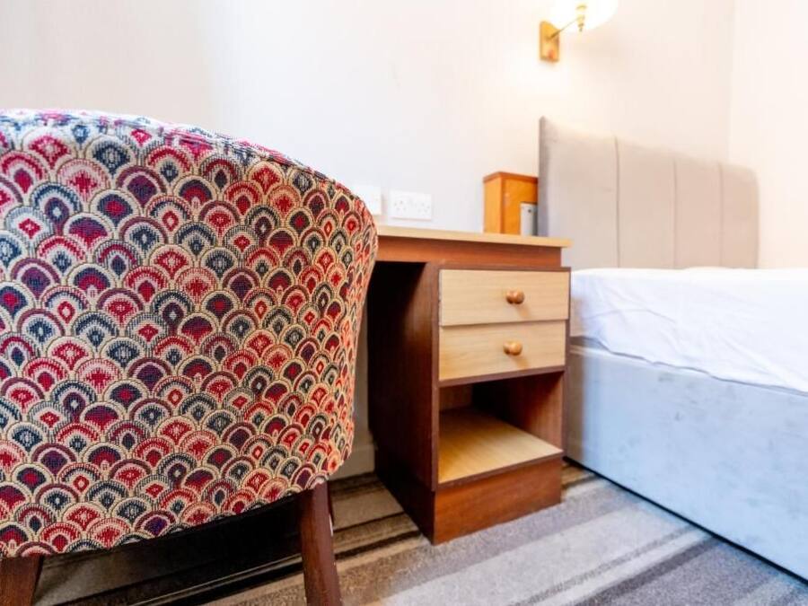 Quarto de hotel com uma cama, uma mesinha de cabeceira de madeira com gavetas e uma cadeira estampada.