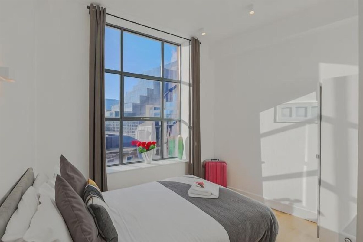 Quarto de hotel com uma janela grande, uma cama com almofadas, um vaso com flores e uma mala.
