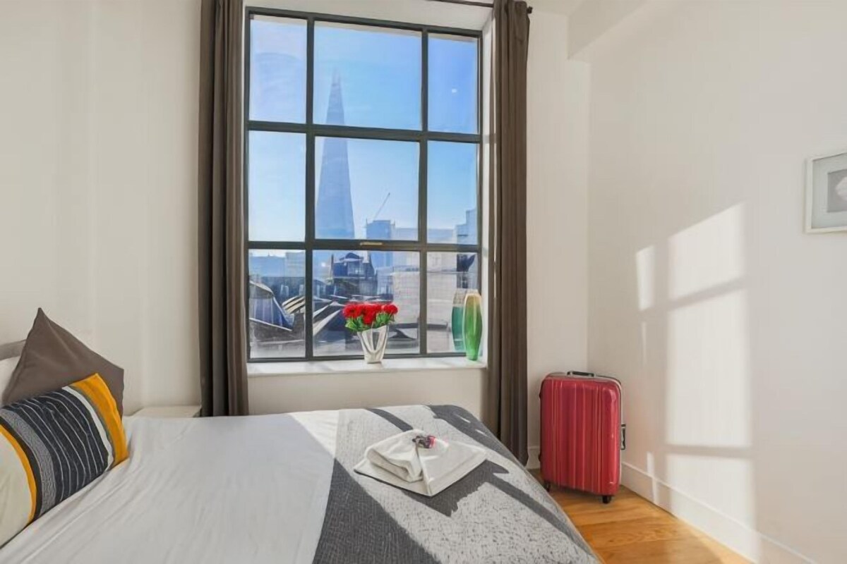 Quarto de hotel com uma janela grande, uma cama com almofadas, uma mala e um vaso com flores.