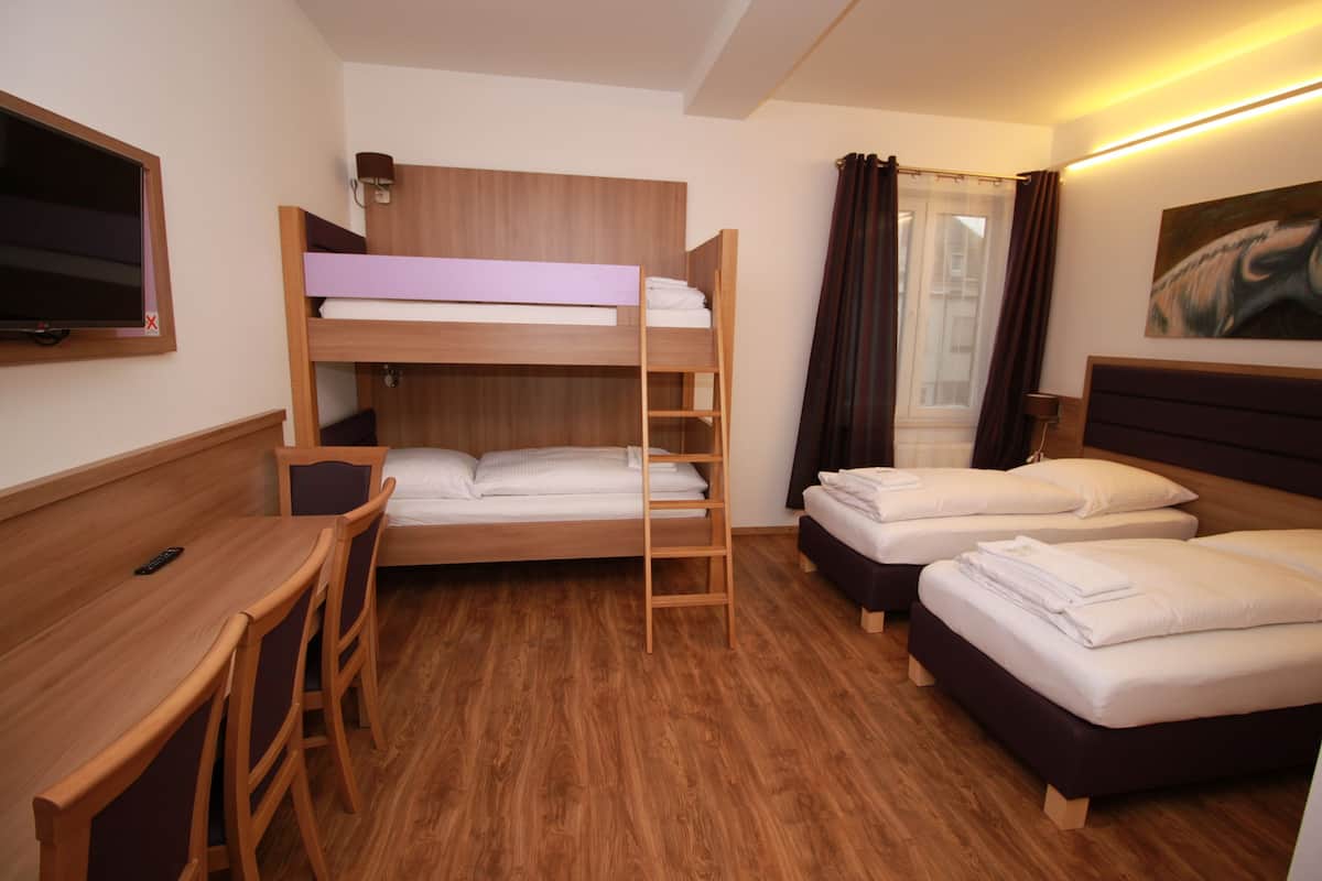 Quarto de hotel com beliches, uma escrivaninha, cadeiras, televisão e um quadro na parede.