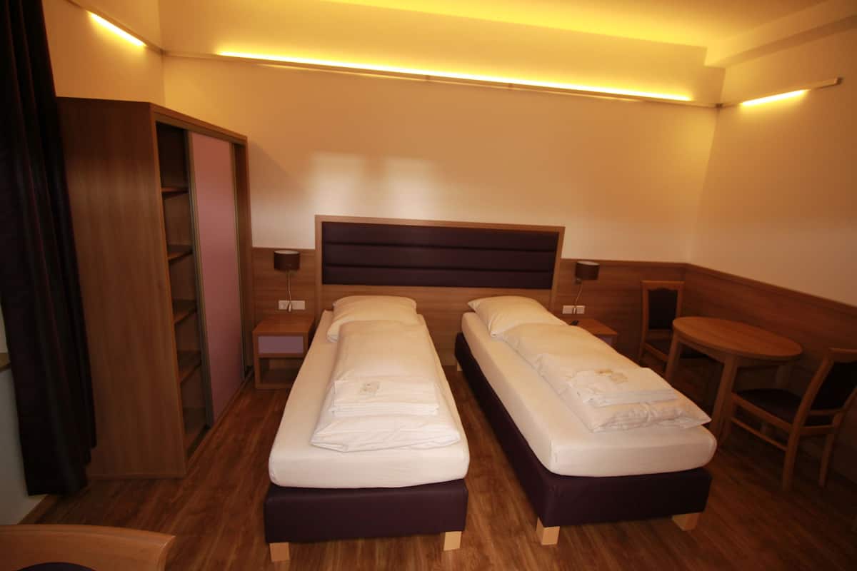 Quarto de hotel com duas camas de solteiro, um guarda-roupa de madeira, uma mesinha e uma cadeira.