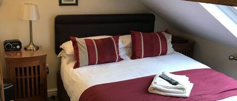 Cama bem arrumada com travesseiro listrado em vermelho e branco, um cobertor branco e um controle remoto sobre ela. Tem uma mesa de cabeceira de madeira com uma luminária e um rádio ao lado da cama.