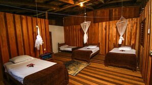 Quarto com paredes e piso de madeira, duas camas com lençóis brancos, um banco de madeira e mosquiteiros pendurados no teto.