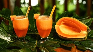 Dois copos de suco de laranja com canudos e metade de uma mamão em uma superfície refletora com folhas verdes.