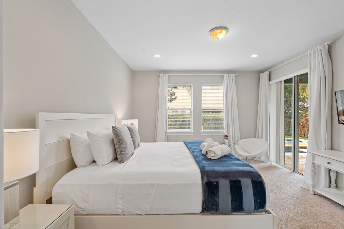 Quarto com uma cama grande, mesa de cabeceira, cadeira e vista para o exterior através de uma janela.