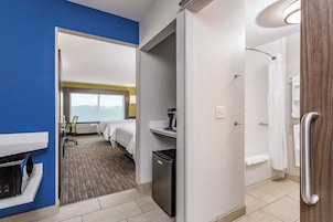Quarto de hotel com cama, escrivaninha, cadeira, lixeira pequena e chuveiro.