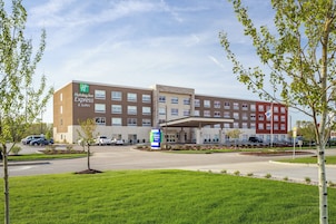 Um hotel Holiday Inn Express & Suites com design moderno, apresentando estacionamento, área de entrada ajardinada e céu claro.