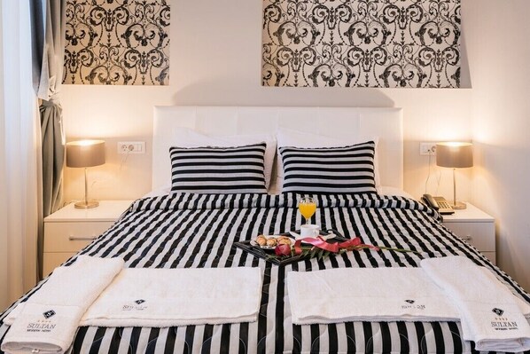 Cama de casal com roupa de cama listrada, duas mesas de cabeceira com abajures, bandeja com itens de café da manhã e toalhas com o logotipo do hotel.