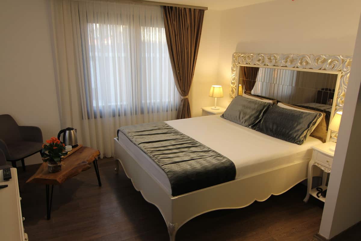 Quarto de hotel com uma cama grande, mesa de cabeceira com abajur, uma cadeira e uma pequena mesa com uma planta.