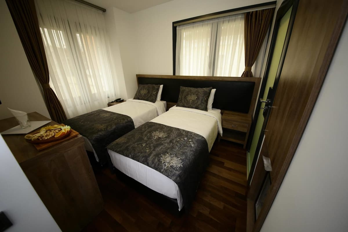 Quarto de hotel com duas camas, cabeceira de madeira, mesa de cabeceira com um prato de comida e janela com cortinas.
