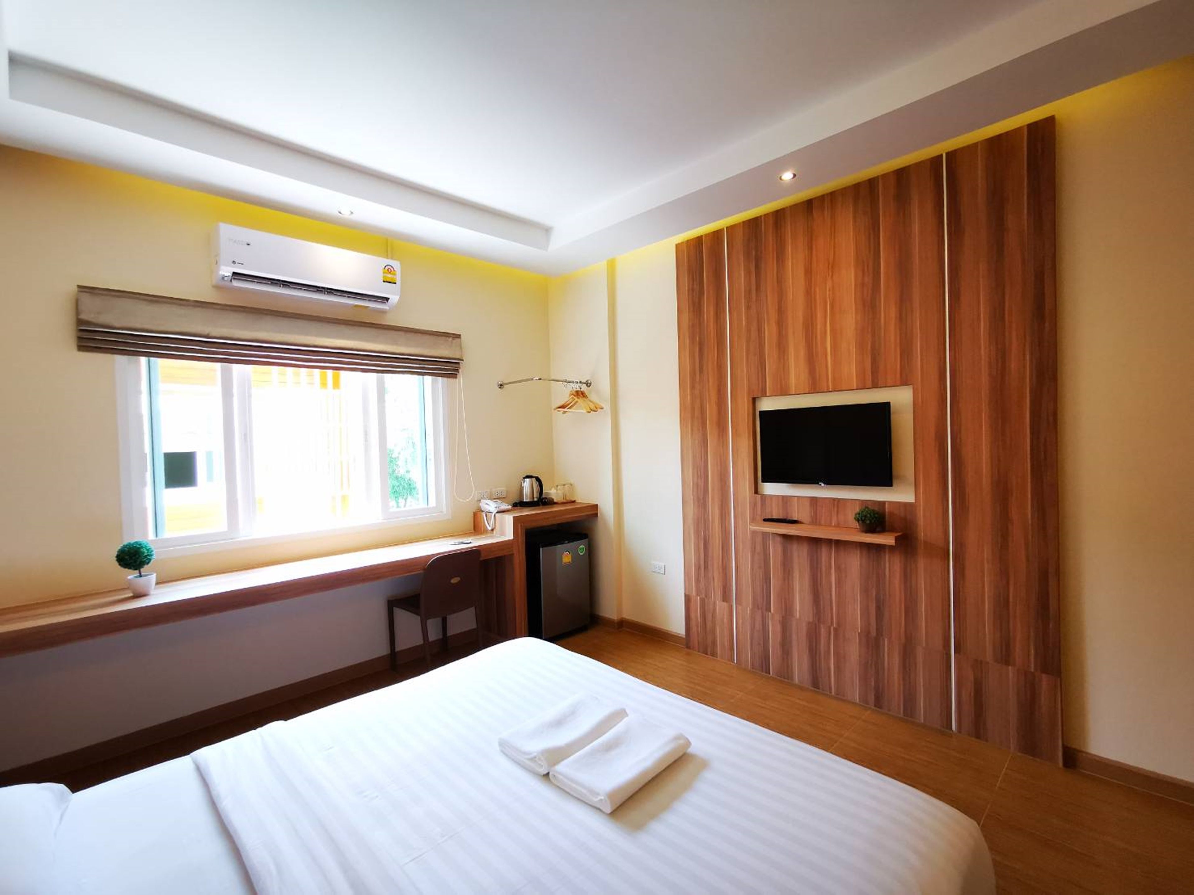 Quarto de hotel com uma cama, uma escrivaninha com cadeira, uma pequena cozinha e televisão fixada numa parede de painel de madeira, além de uma janela com vista para o exterior.