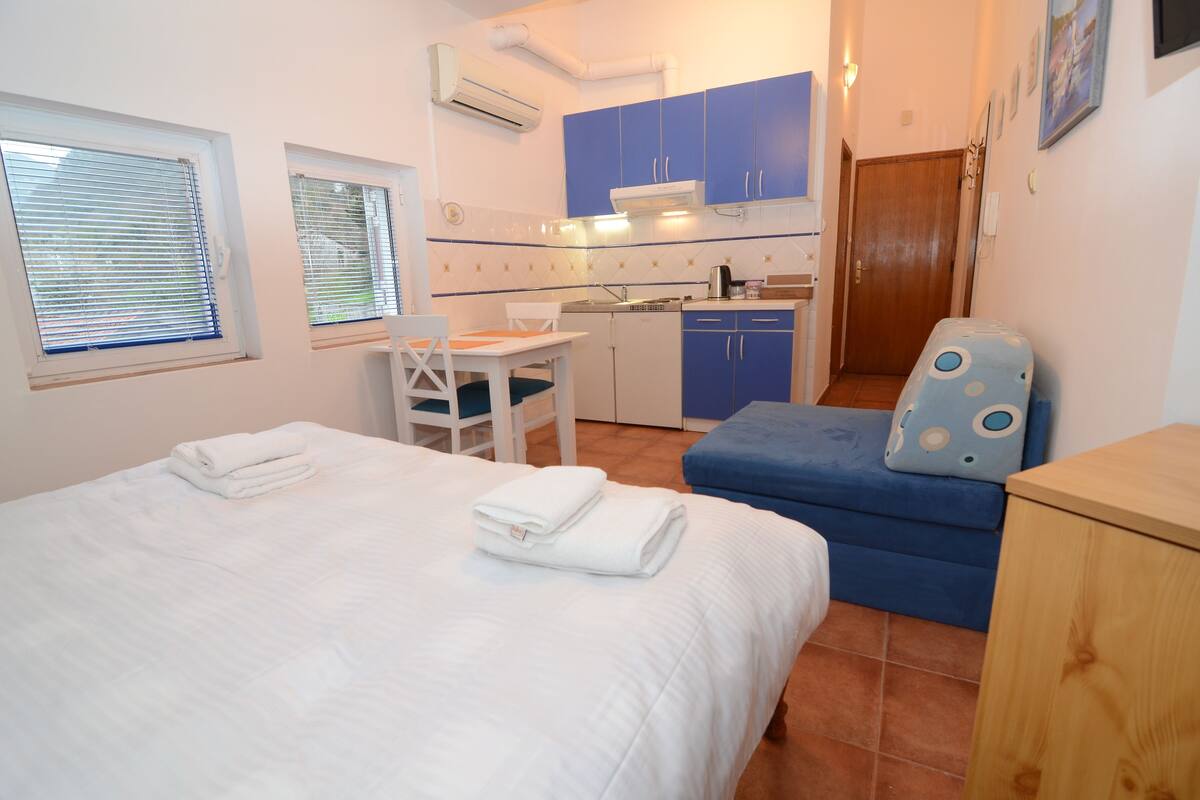Quarto compacto com cama, pequena escrivaninha, cozinha compacta e sofá azul.