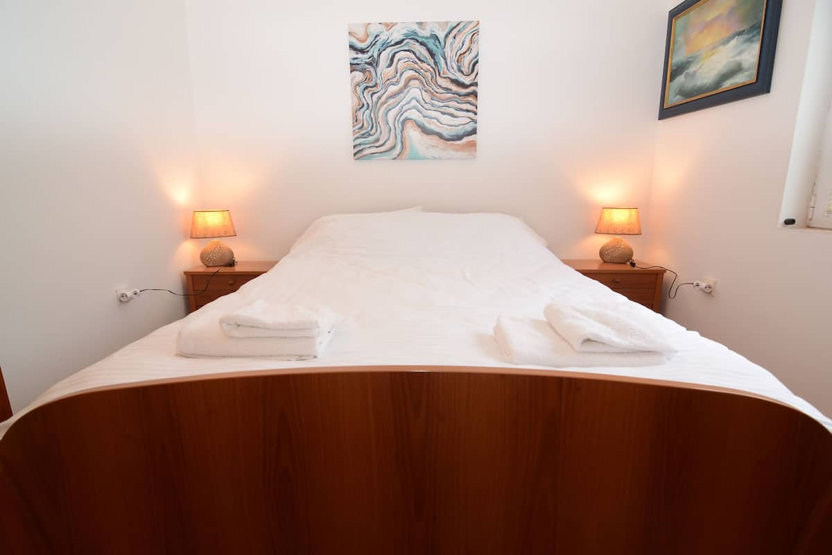Uma cama bem arrumada com duas luminárias de cabeceira, uma obra de arte na parede e uma janela com persianas.