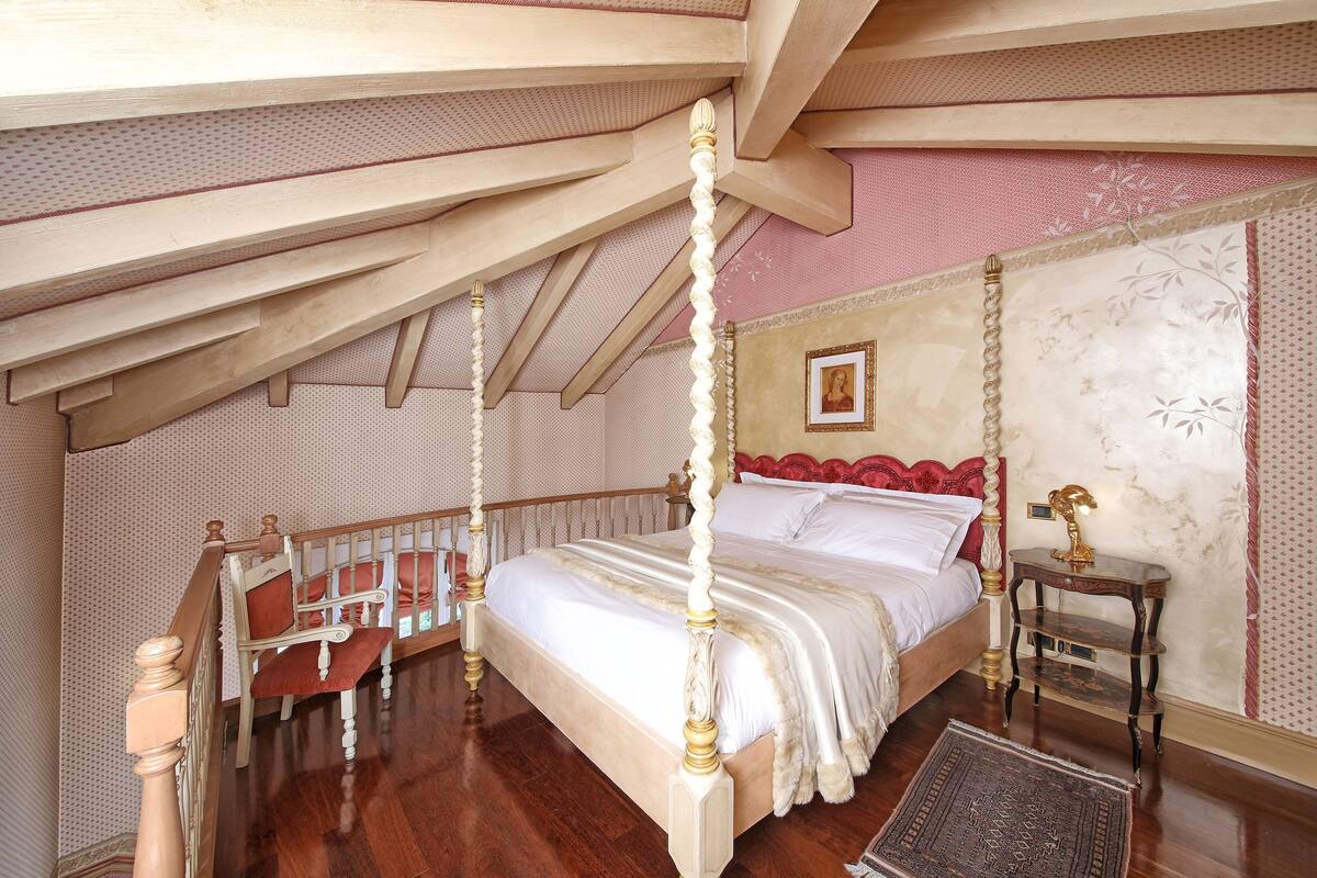 Uma cama com dossel, estrutura de madeira e roupa de cama branca, em um quarto com piso de madeira e teto inclinado.