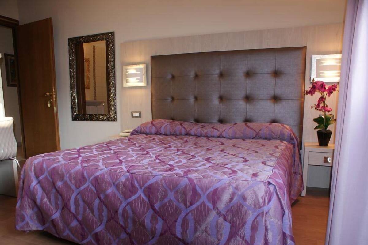 Quarto com cama grande, cabeceira, mesa de cabeceira, espelho e planta em vaso.