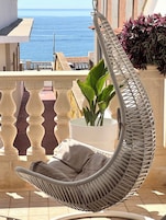 Uma varanda com uma cadeira suspensa, um vaso de planta e vista para o mar.
