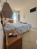 Quarto com cama, mesa de cabeceira com controle remoto, luminária pendente, guarda-roupa e vista para o exterior.