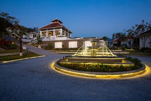 Uma fonte bem iluminada no centro de uma entrada circular, rodeada por canteiros de flores e um edifício com arquitetura tradicional.