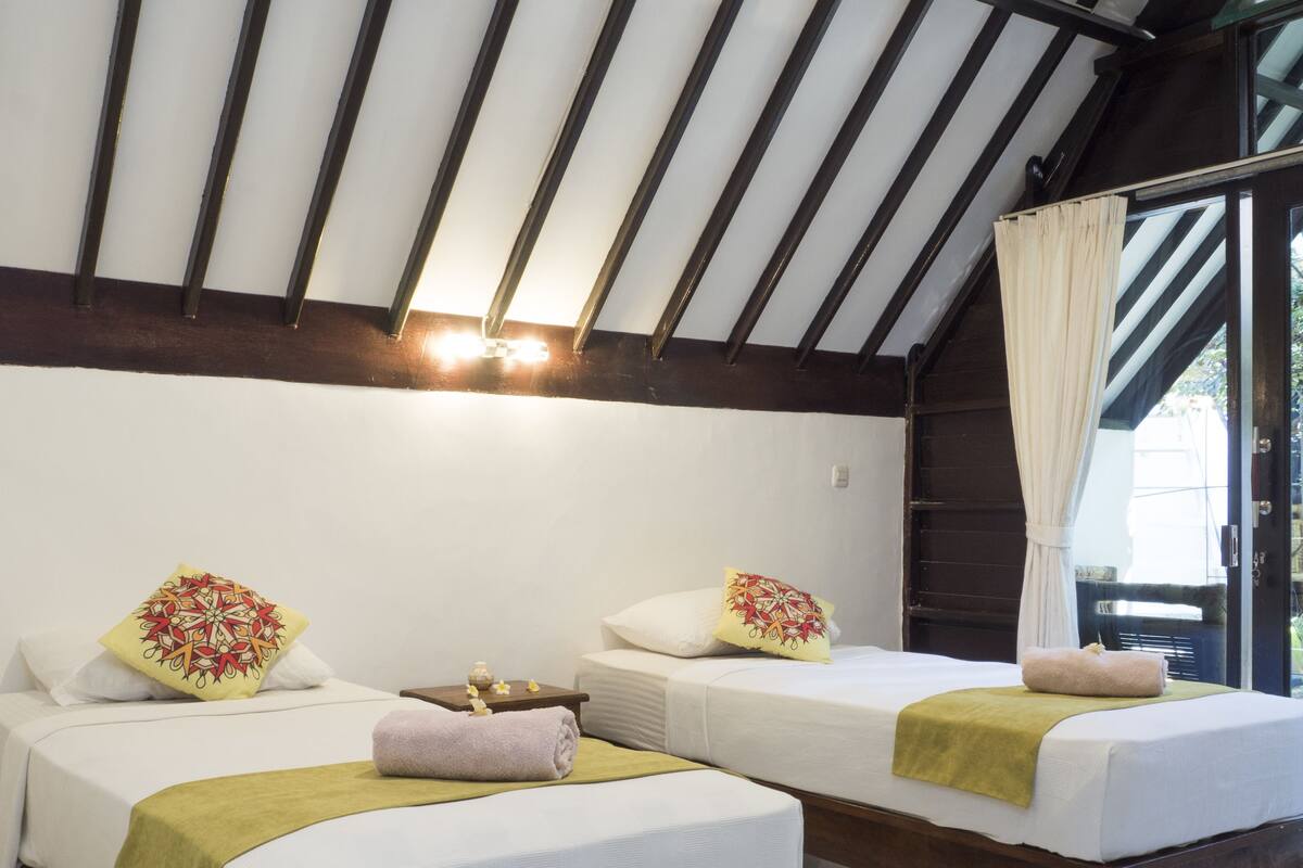 Quarto com duas camas, cada uma com estrutura de madeira e roupa de cama branca. Há almofadas decorativas e uma pequena mesa com luminária entre as camas. O quarto tem teto inclinado com vigas de madeira e uma janela com cortinas.