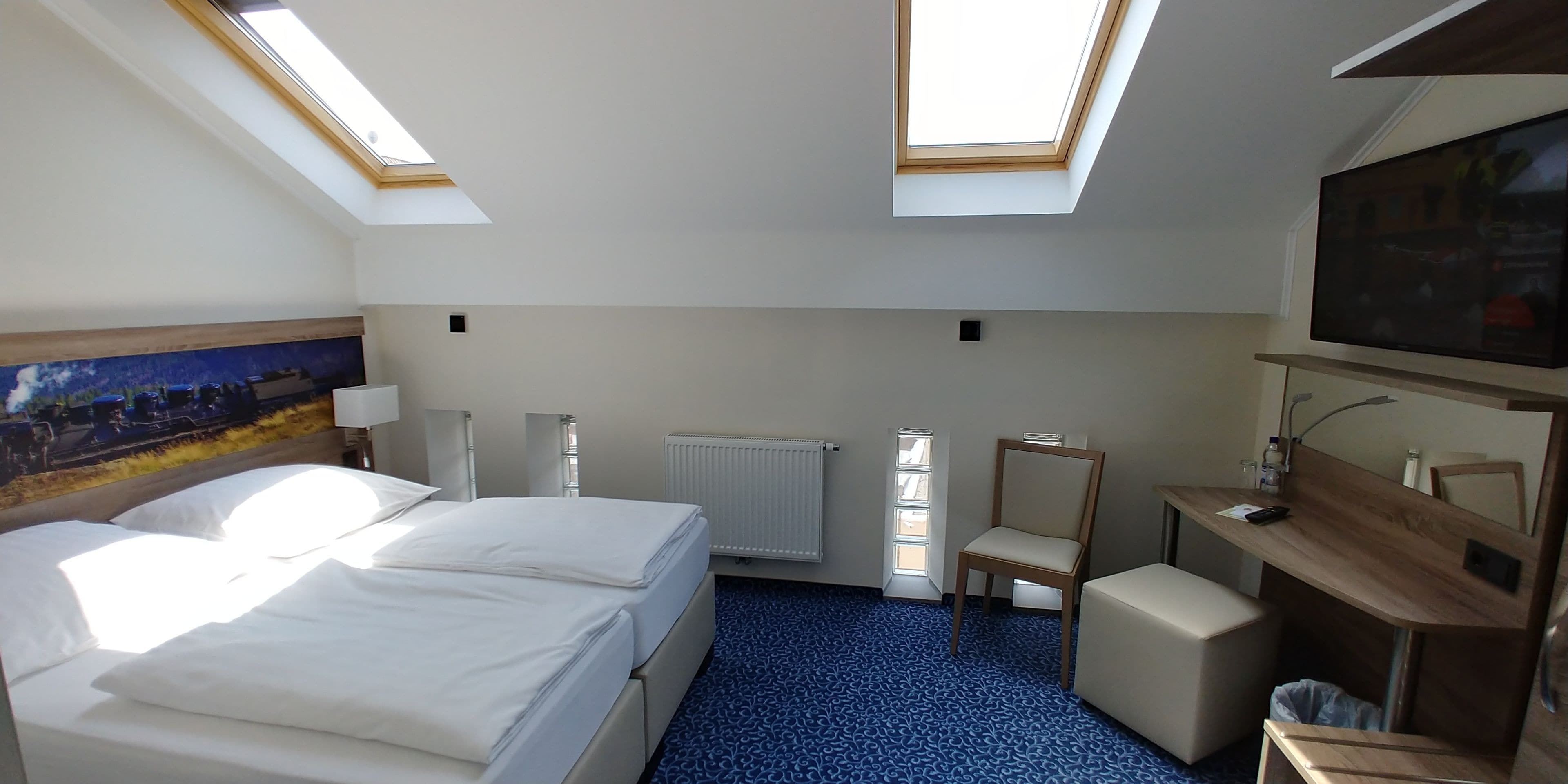 Quarto de hotel com cama, escrivaninha, cadeira, televisão, radiador e claraboias.