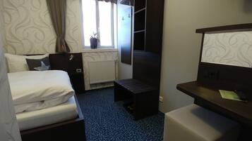 Quarto de hotel com cama, escrivaninha, cadeira, radiador e janela com cortinas.