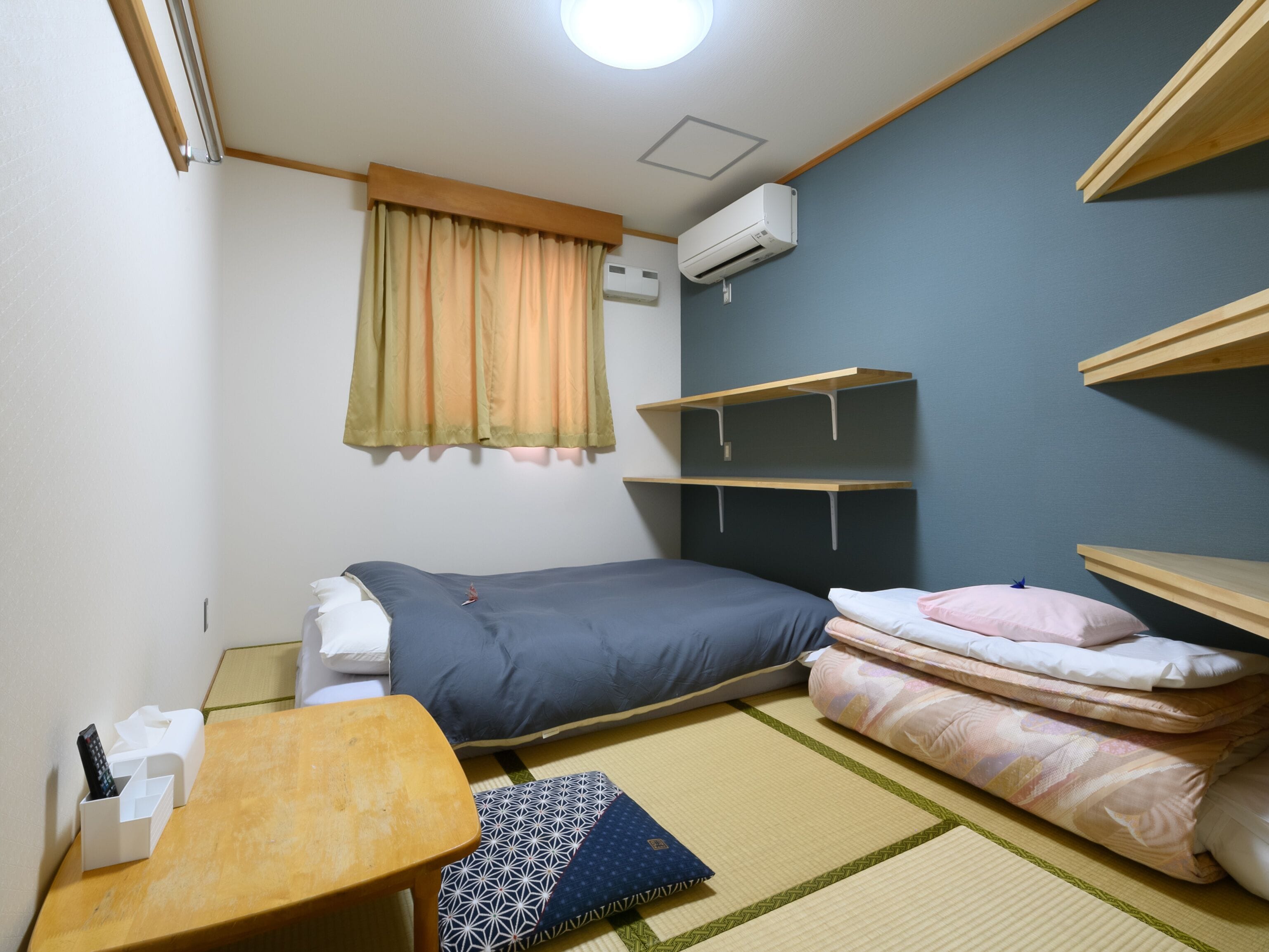 Quarto pequeno e bem organizado, com cama, mesa de cabeceira, cadeira e estante.