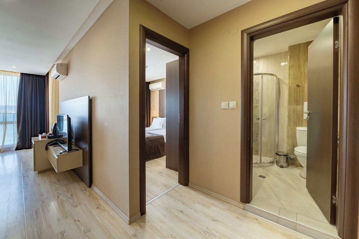 Quarto de hotel com cama, escrivaninha com televisão, área de chuveiro e vista para o exterior.
